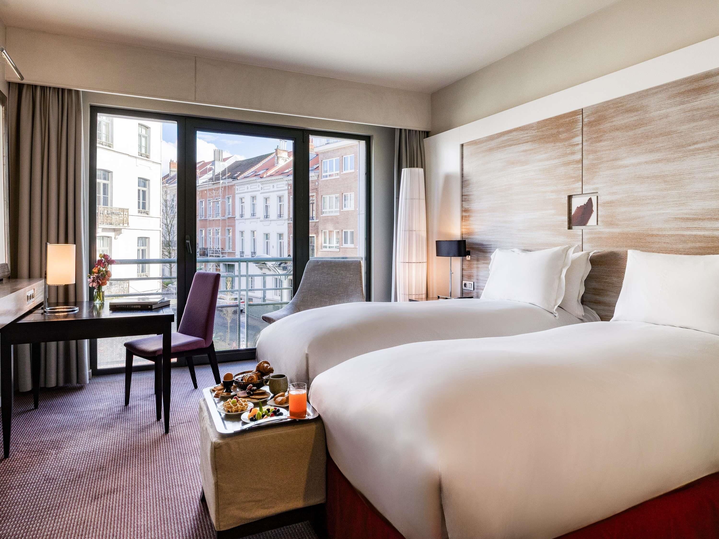 Quarto de hotel com cama, escrivaninha, cadeira, mesa pequena com itens de café da manhã e vista para edifícios por grandes janelas.