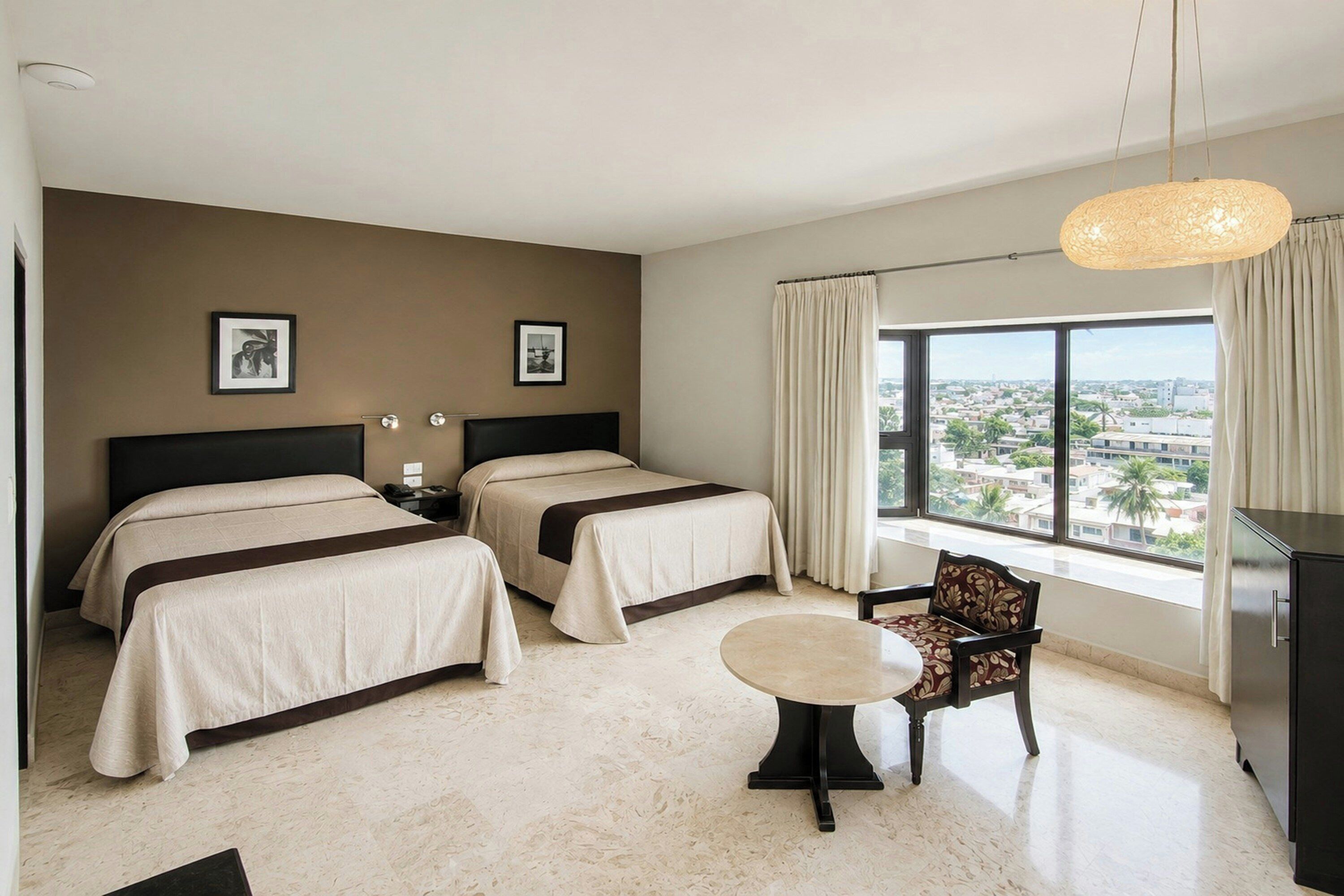 Quarto de hotel com duas camas, uma mesa pequena, uma cadeira, escrivaninha e uma janela grande com vista para a cidade.