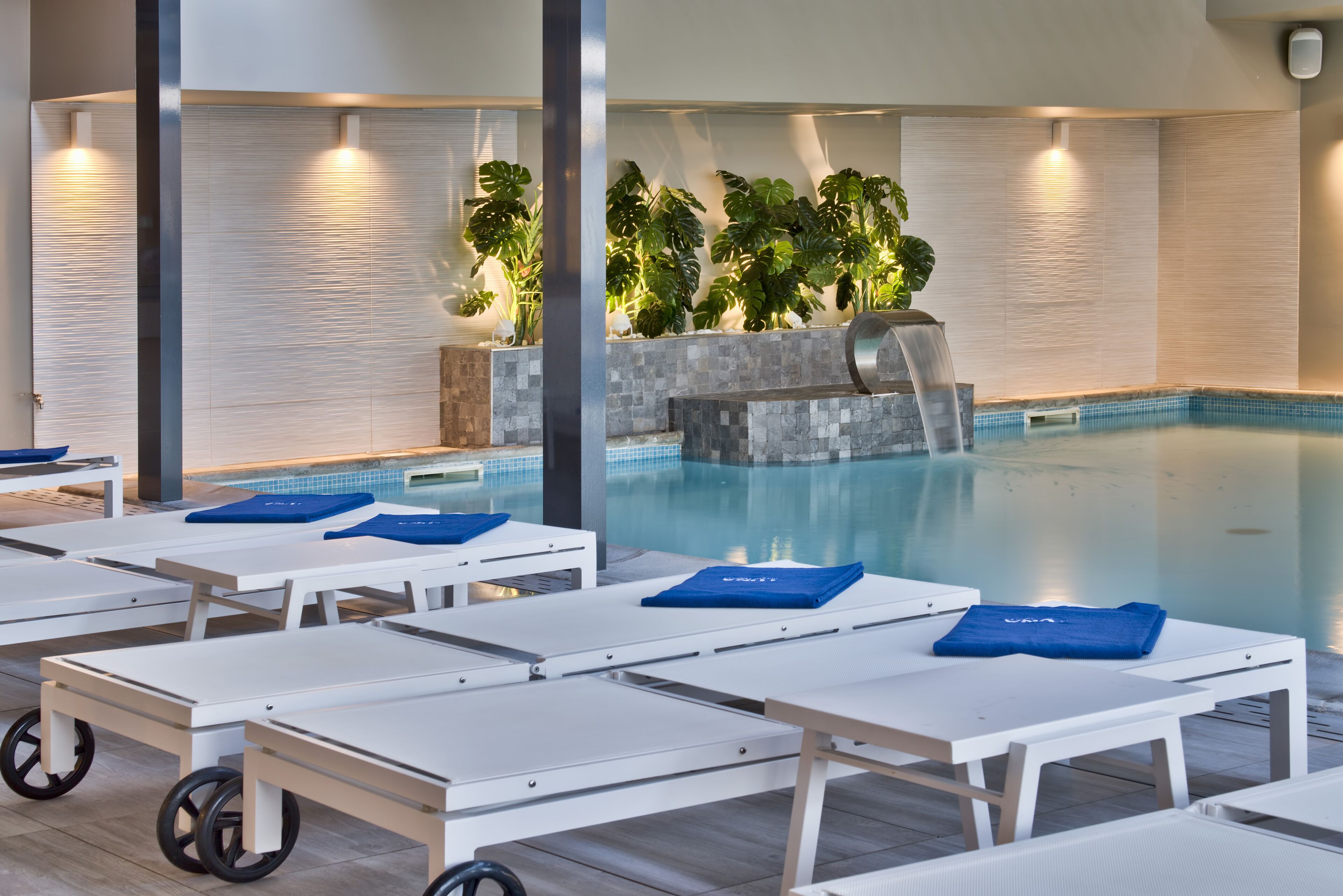 Uma área junto à piscina com espreguiçadeiras, uma cascata e plantas em vasos.