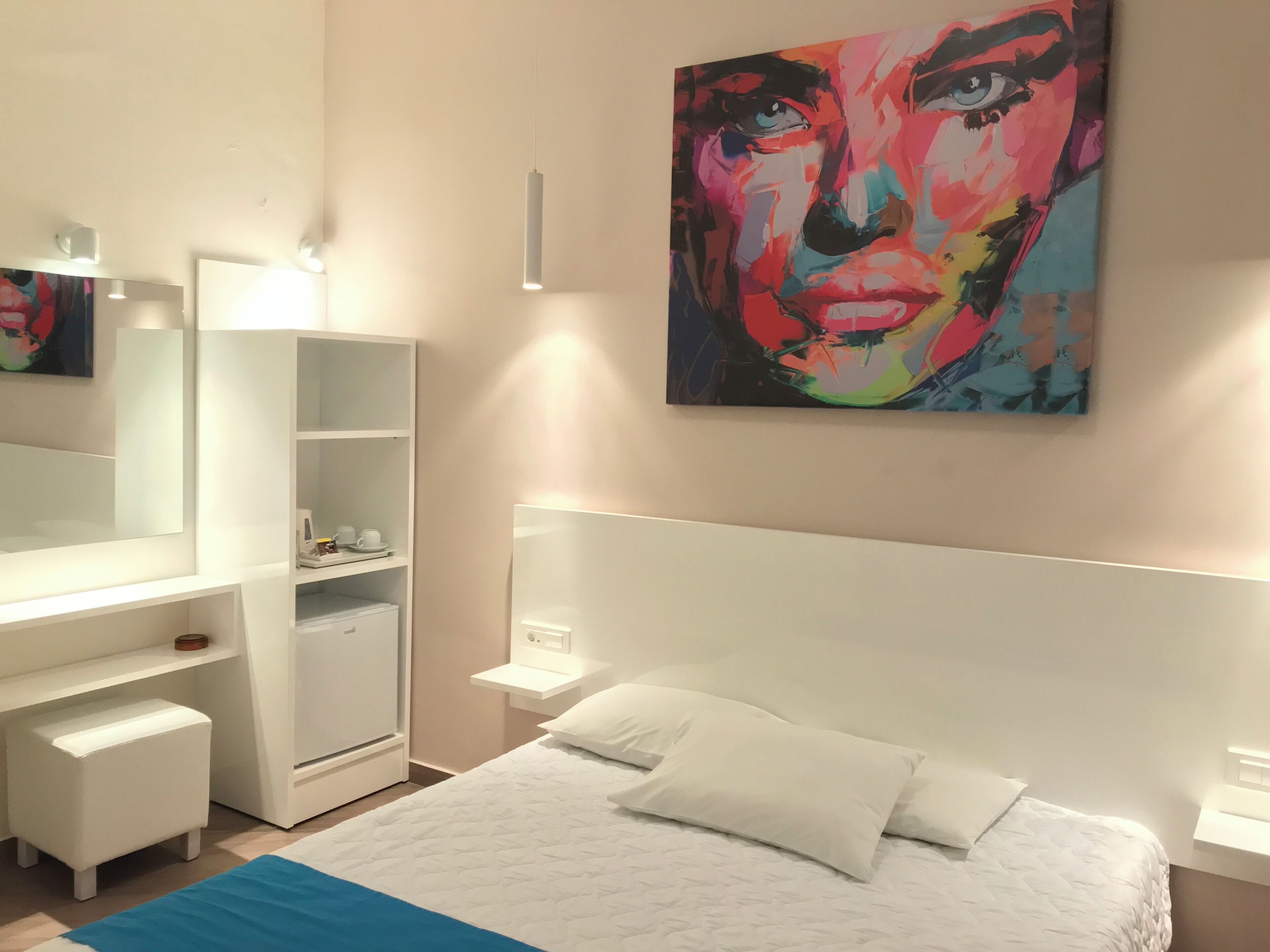 Quarto de hotel com cama, mesa de cabeceira, armário, espelho e um quadro colorido na parede.