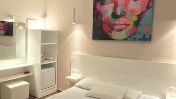 Quarto de hotel com cama, mesa de cabeceira, armário, espelho e um quadro colorido na parede.