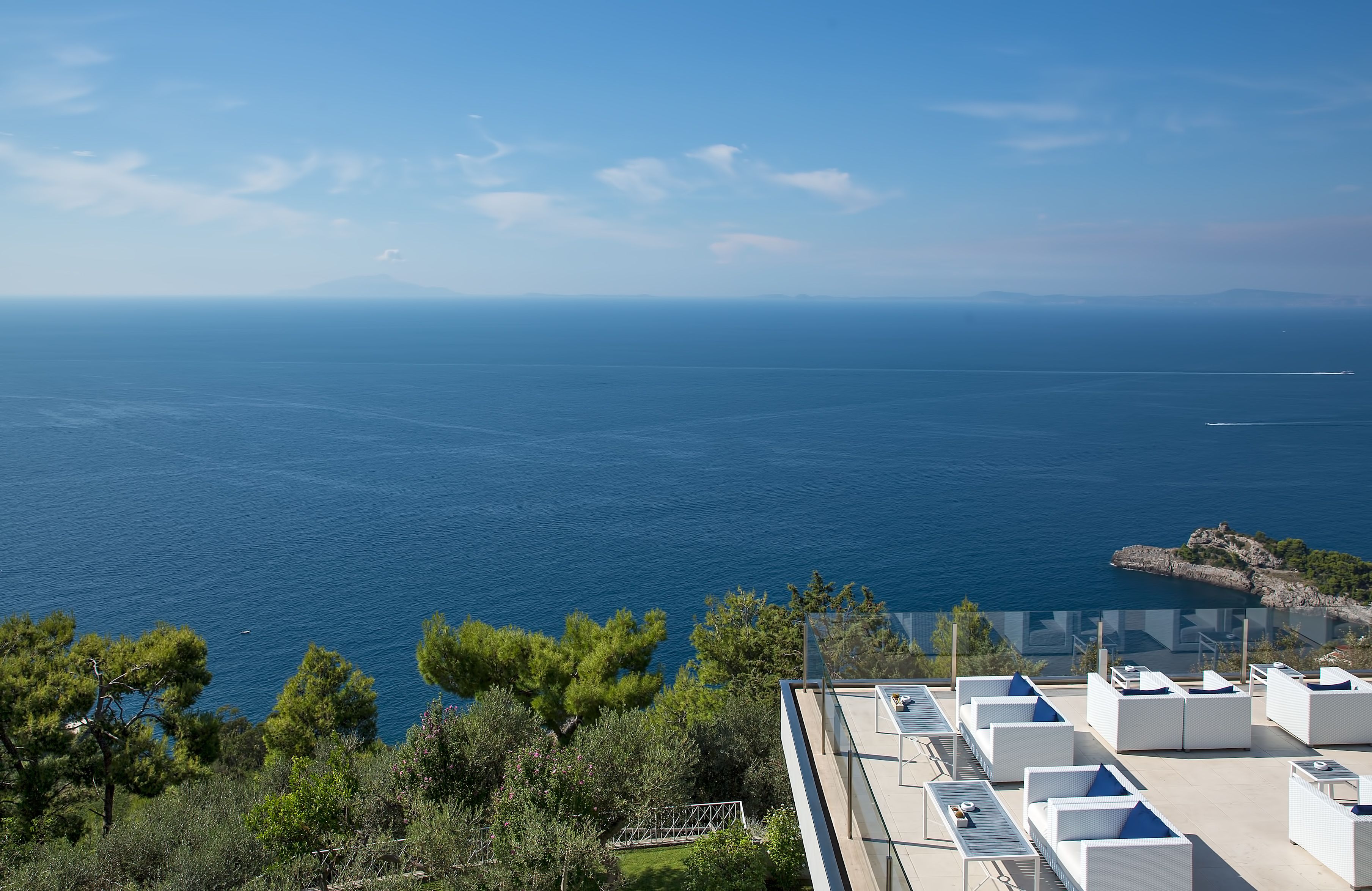 Um terraço à beira-mar com mobiliário de exterior branco e vista para o mar e montanhas ao longe.