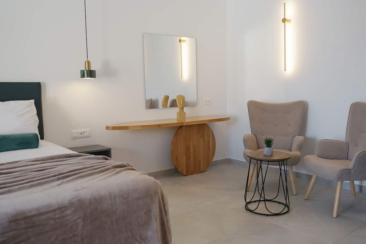 Quarto de hotel moderno com uma cama, uma mesinha redonda com uma planta, duas poltronas, um console de madeira e um espelho com luminária.
