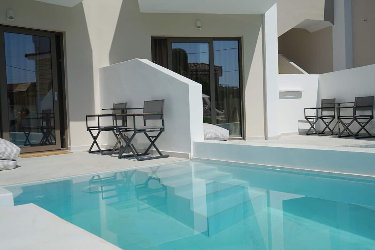 Uma área de piscina moderna com água azul cristalina, uma espreguiçadeira e duas áreas de estar com mesas e cadeiras.