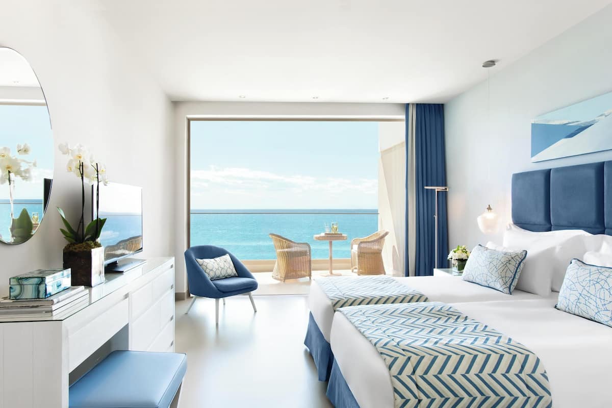 Quarto de hotel moderno com uma cama grande, uma mesa com espelho e vista para o oceano.