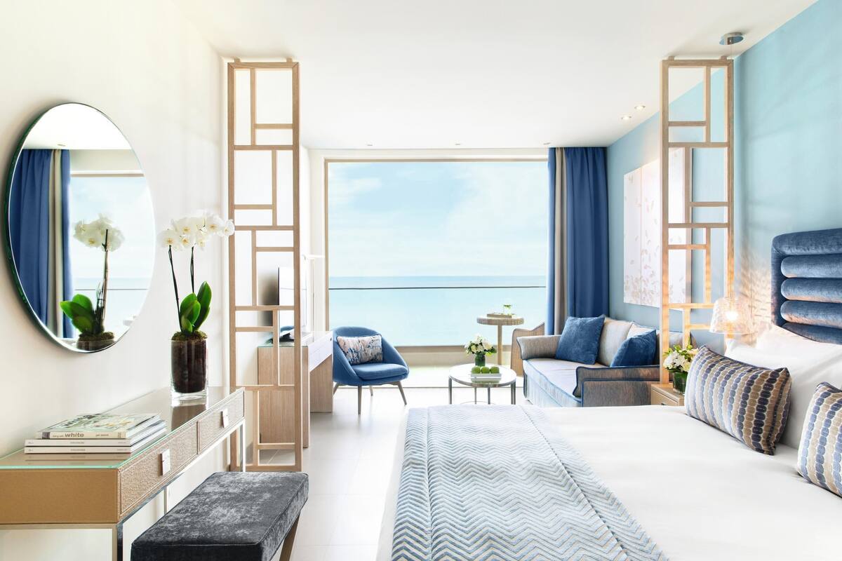 Quarto de hotel moderno com uma cama grande, área de estar com vista para o mar, uma mesinha com um vaso de flores e um espelho grande.