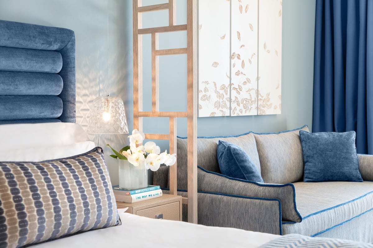 Quarto de hotel moderno com cama, sofá, mesa de cabeceira com livros e flores, e um painel decorativo na parede.