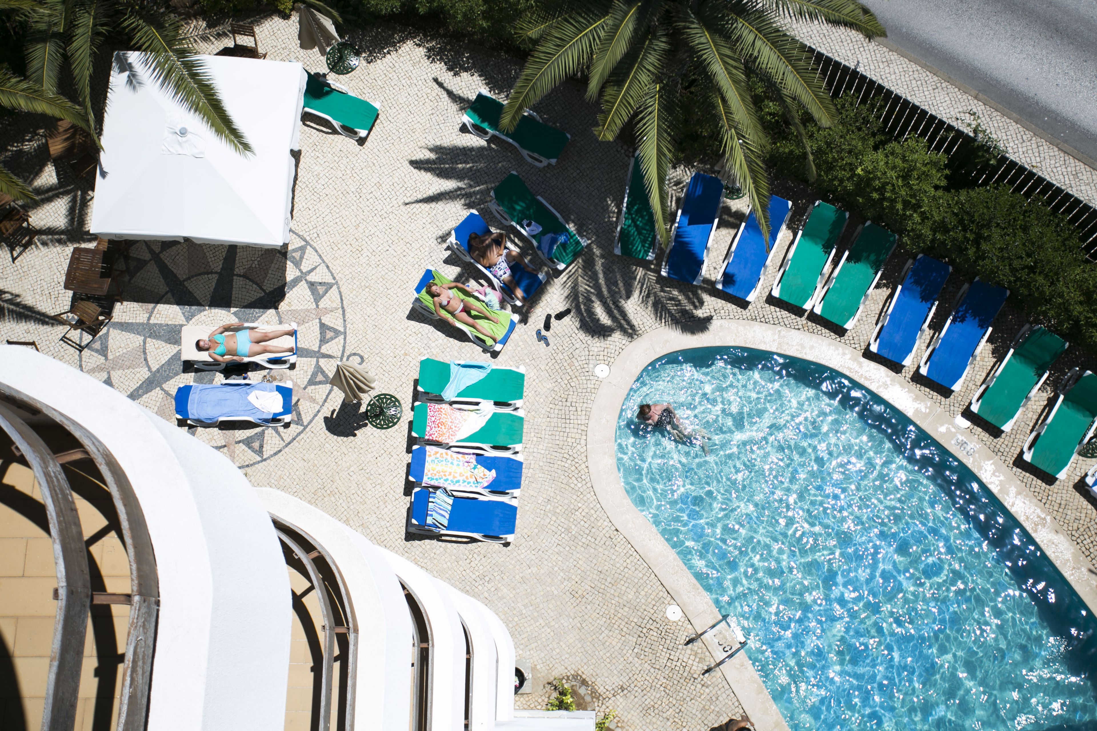 Uma área de piscina no terraço com espreguiçadeiras e guarda-sóis, cercada por palmeiras.