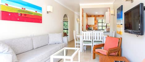 Uma sala de estar com sofá cinzento, mesa de centro de vidro, mesa de jantar de madeira com cadeiras, televisão presa à parede e uma cozinha ao fundo.