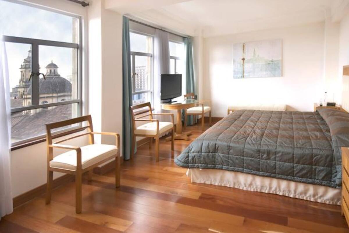 Quarto de hotel com cama, escrivaninha, cadeira, televisão e janela com vista para a cidade.