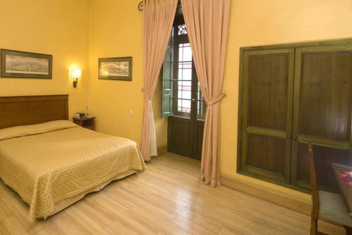 Quarto com cama de madeira, luminárias de cabeceira, mesa de cabeceira, janela com cortinas e guarda-roupa de madeira.