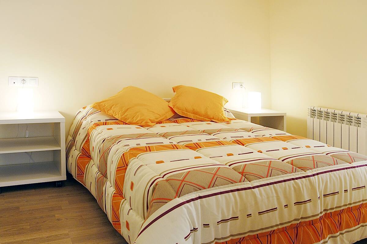 Cama bem arrumada com colcha estampada e dois travesseiros, com mesas de cabeceira com abajures de cada lado, em um quarto com piso de madeira e radiador na parede.