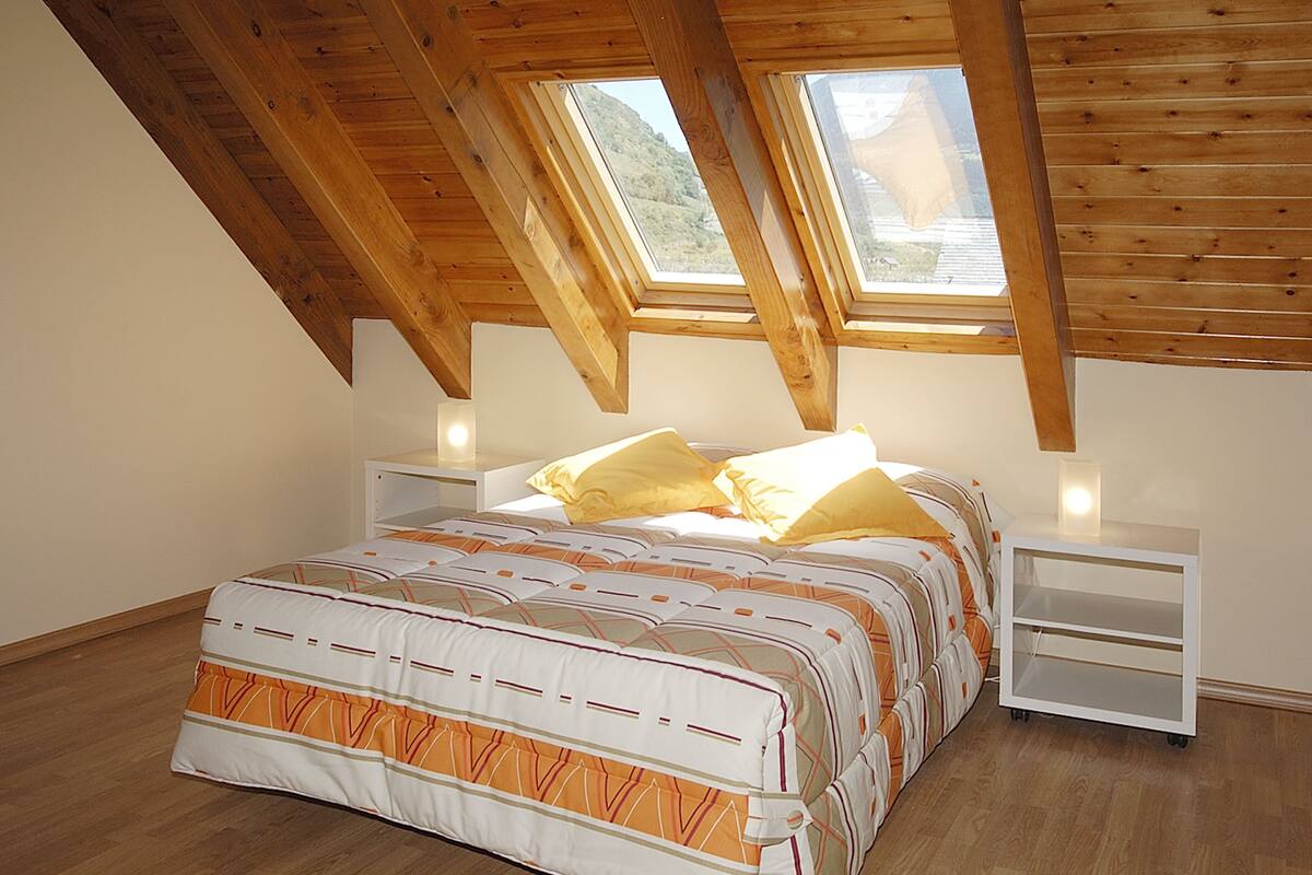 Quarto com teto inclinado, cama com roupa de cama estampada, duas mesas de cabeceira e um claraboia.