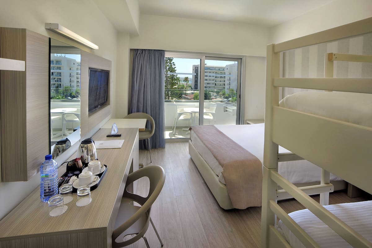 Quarto de hotel com beliche, escrivaninha com cadeira, televisão, janela grande com vista para edifícios e varanda.