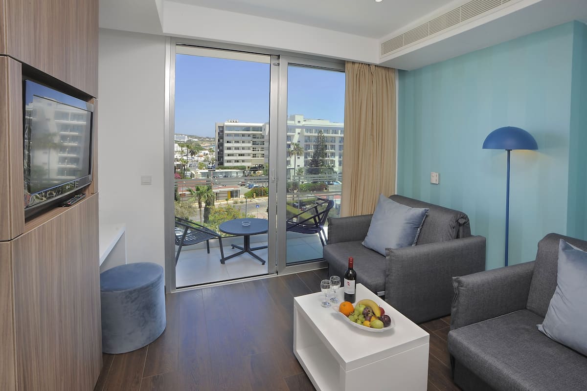 Sala de estar moderna com televisão de tela plana, sofás cinzas, uma mesa de centro com frutas e uma garrafa, e vista para a paisagem urbana através de grandes portas de vidro.