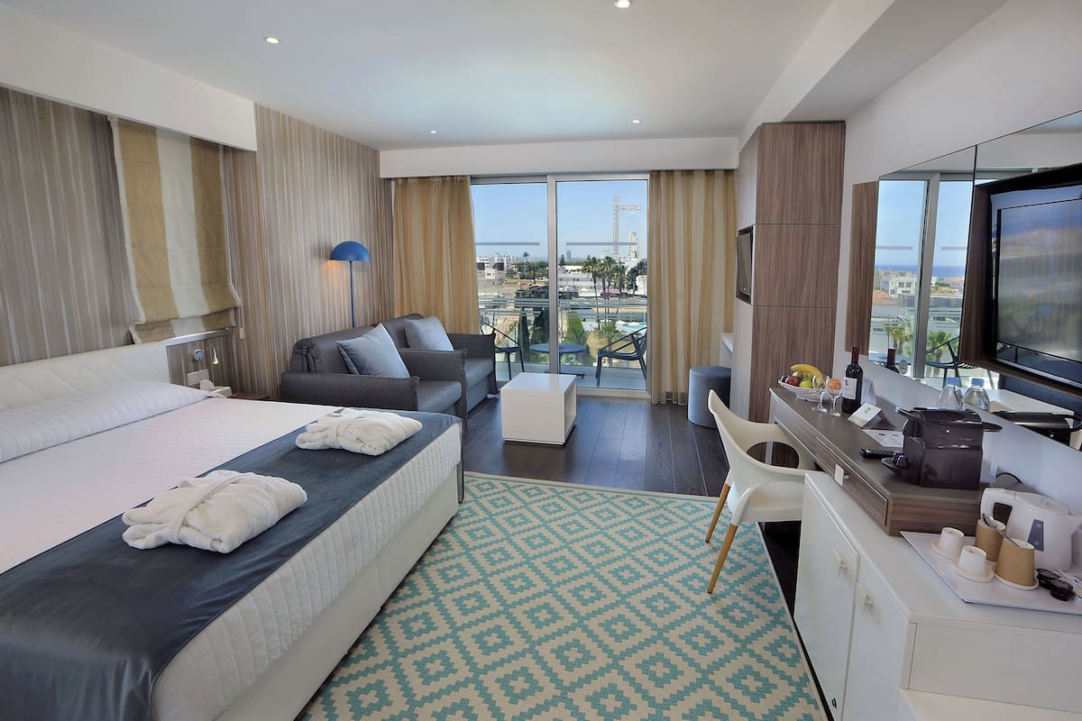 Quarto de hotel com cama, sofá, escrivaninha com televisão, varanda e uma grande janela.