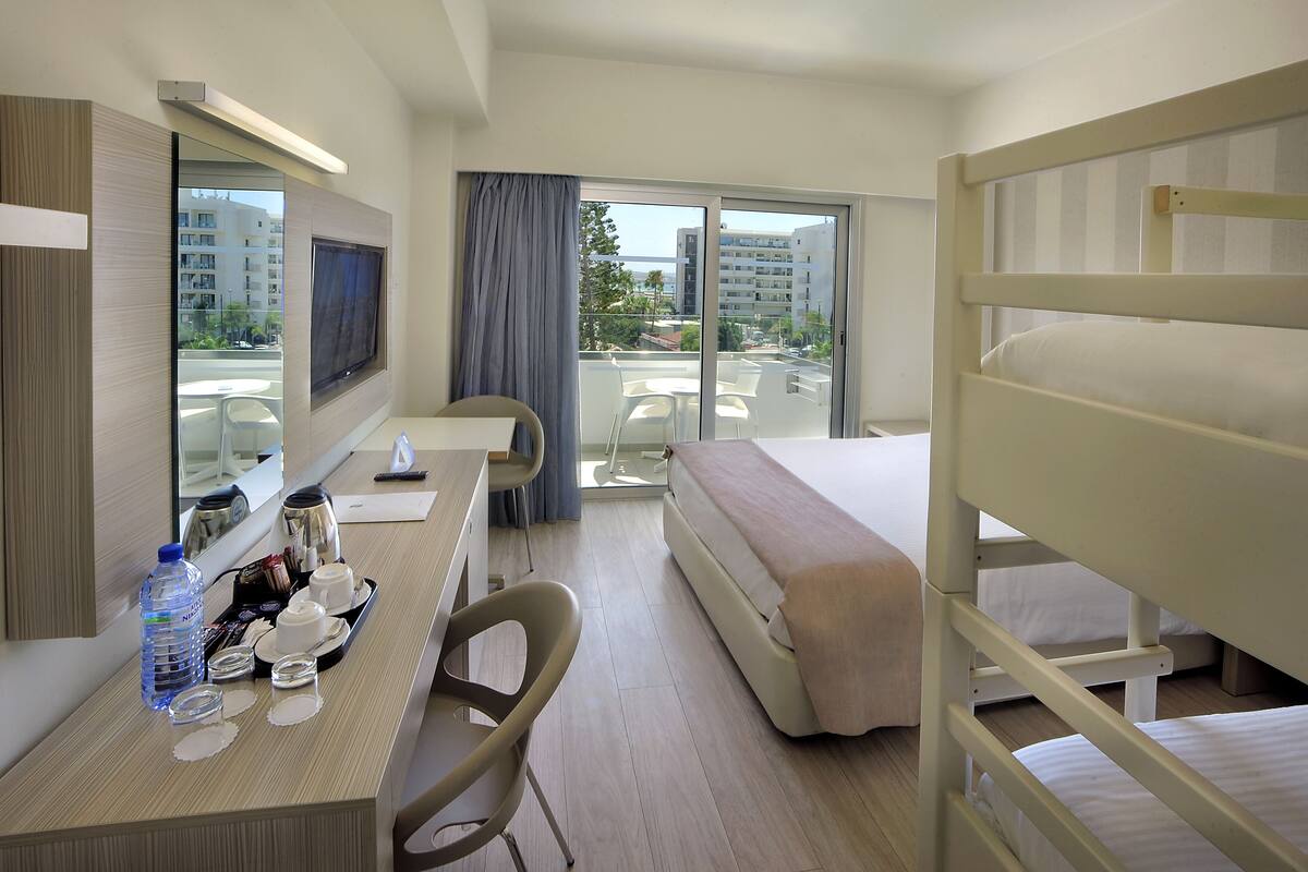 Quarto de hotel com beliche, escrivaninha com cadeira, televisão e varanda com vista para edifícios.