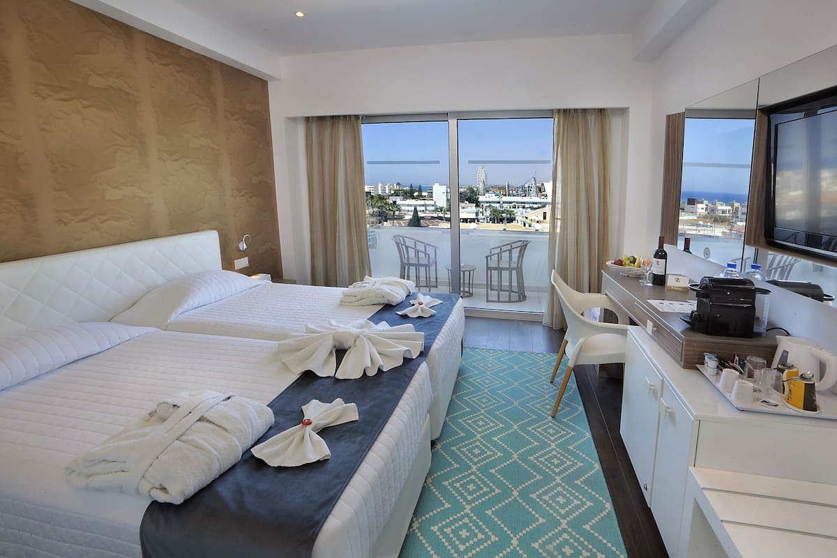 Quarto de hotel com duas camas, uma escrivaninha com televisão, varanda com cadeiras e vista para a cidade.