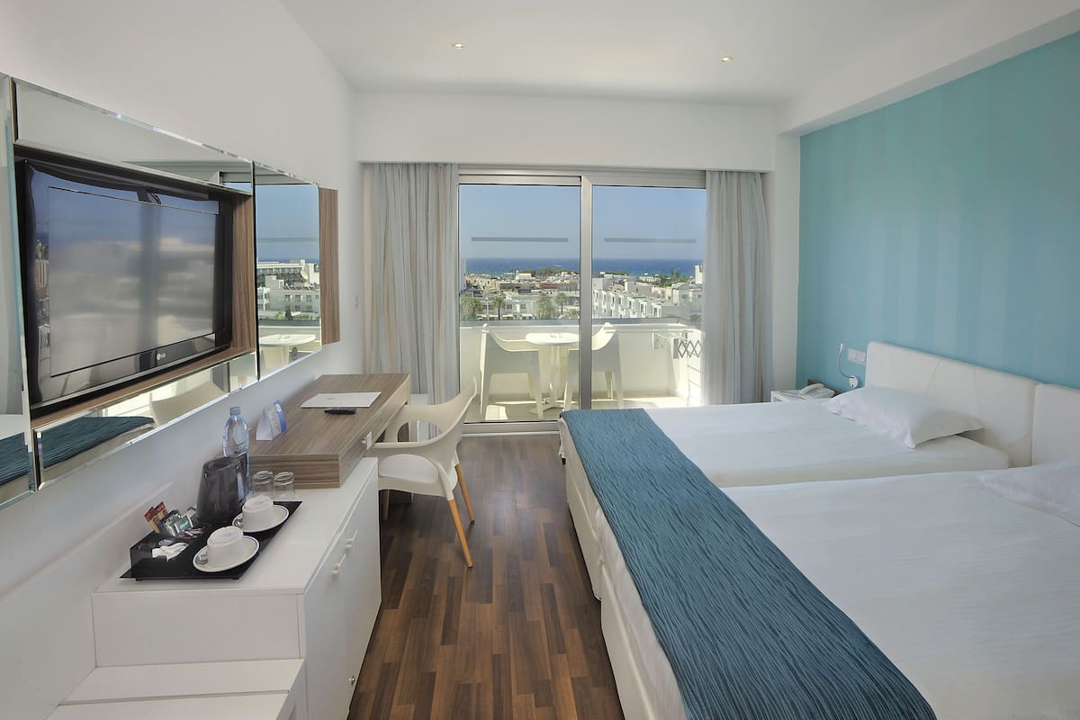 Quarto de hotel com cama, escrivaninha, cadeira, televisão e vista para a cidade e mar.