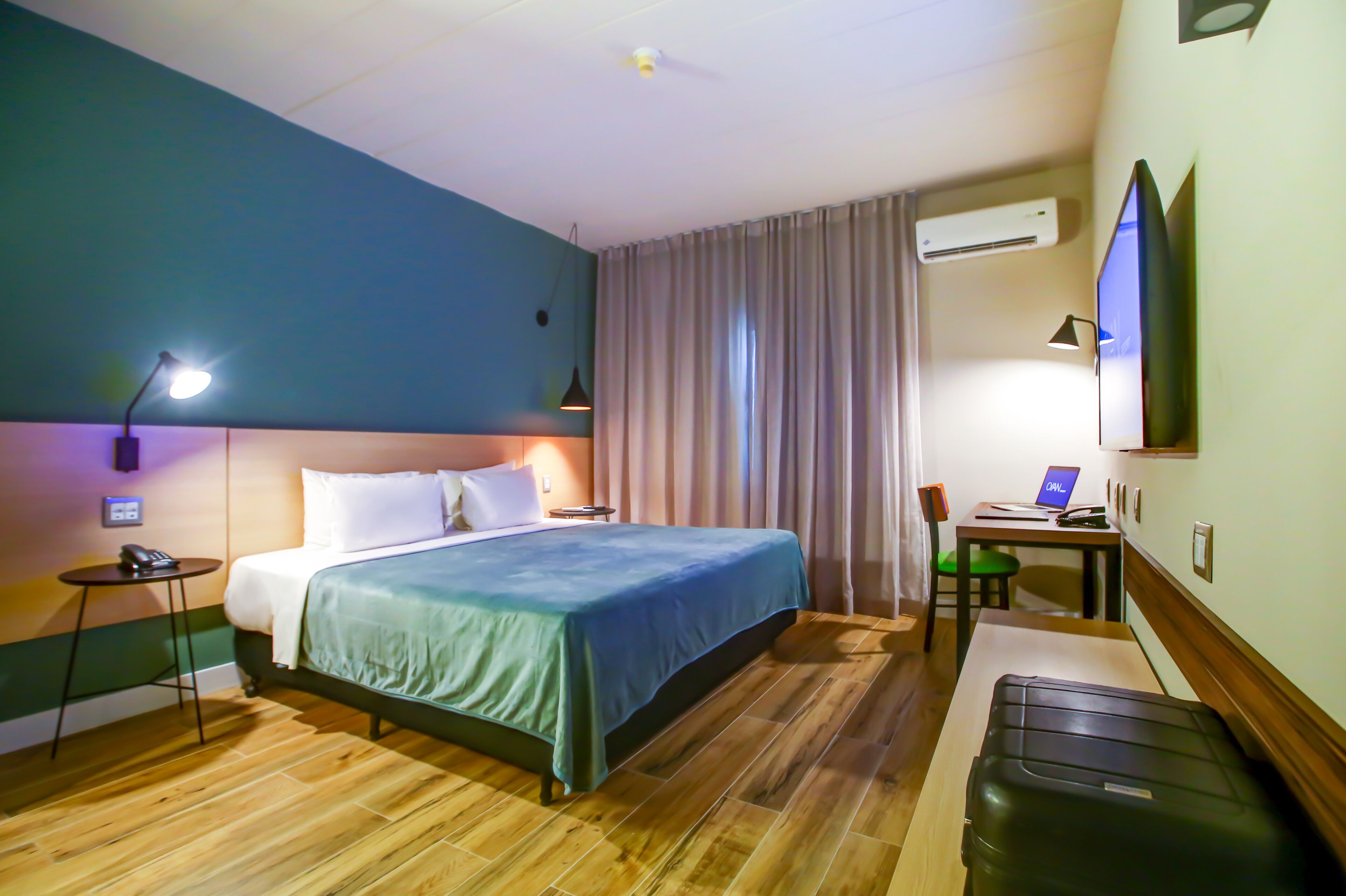 Quarto de hotel com cama, escrivaninha, laptop, cadeira, mala e televisão na parede.