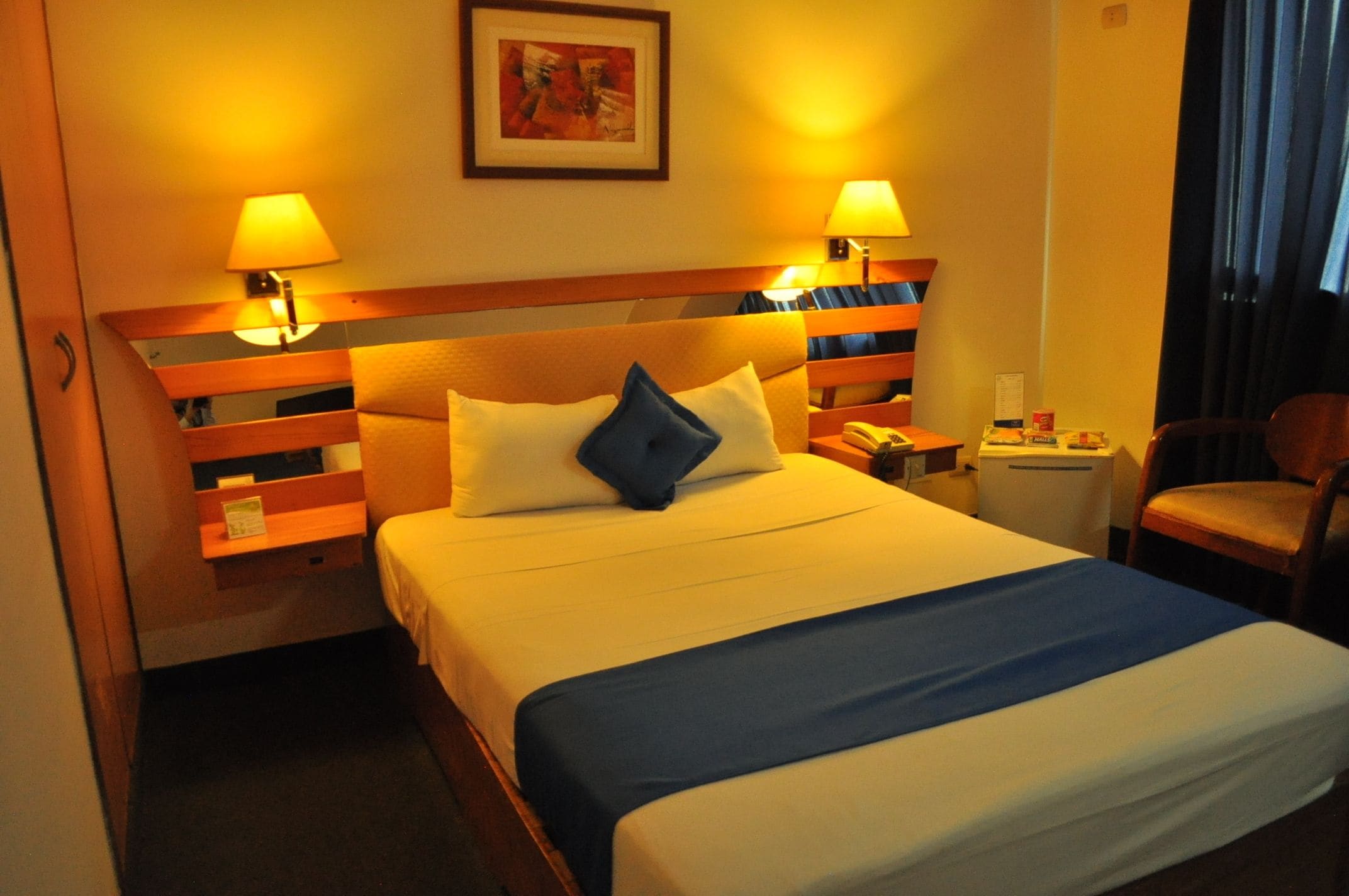 Quarto de hotel com uma cama, duas luminárias de cabeceira, uma poltrona, mesa de cabeceira e um quadro na parede.