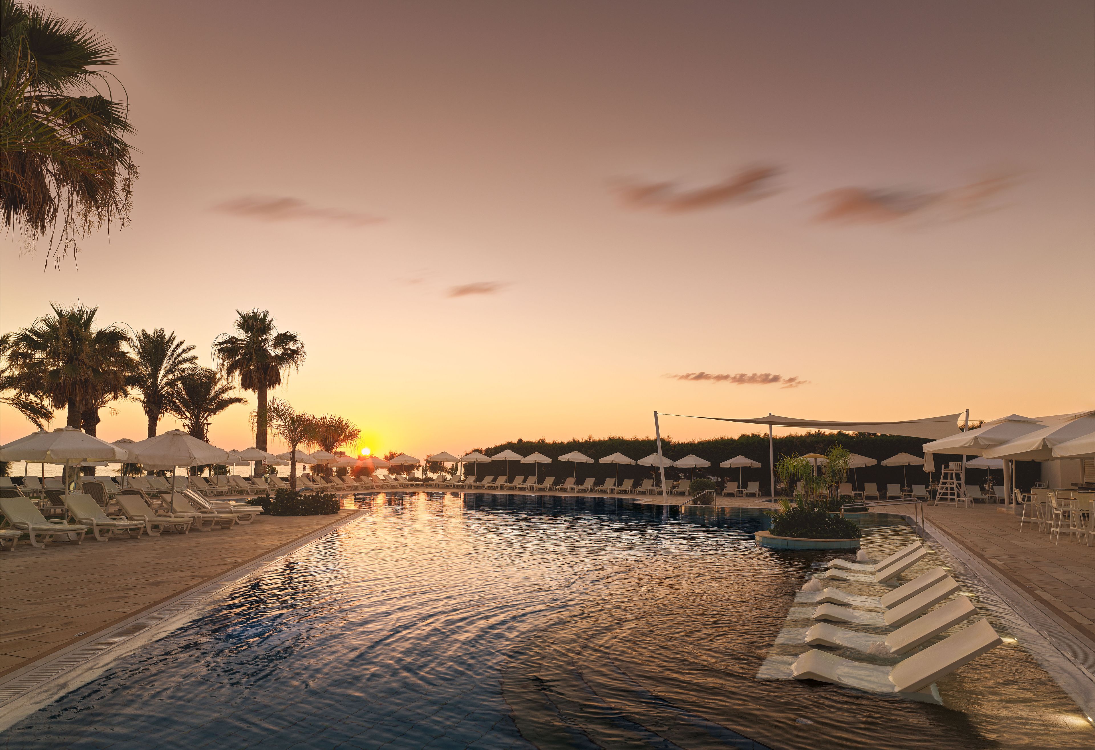 Área de piscina com espreguiçadeiras e guarda-sóis, céu claro e pôr do sol.