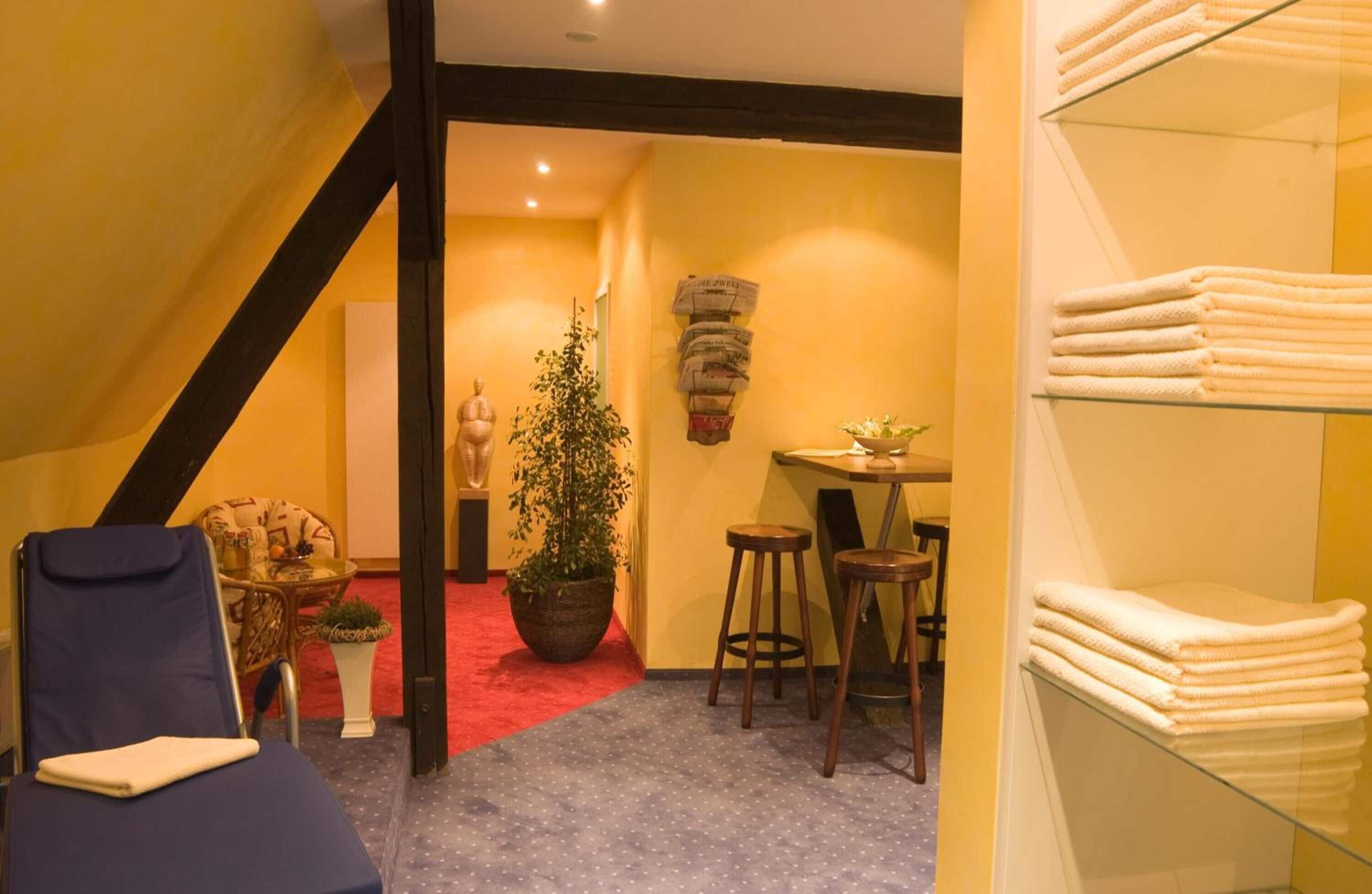 Um hall de hotel com carpete vermelho, uma área de estar com uma cadeira azul, uma pequena mesa com banquinhos, uma planta em vaso e uma estante com toalhas dobradas.
