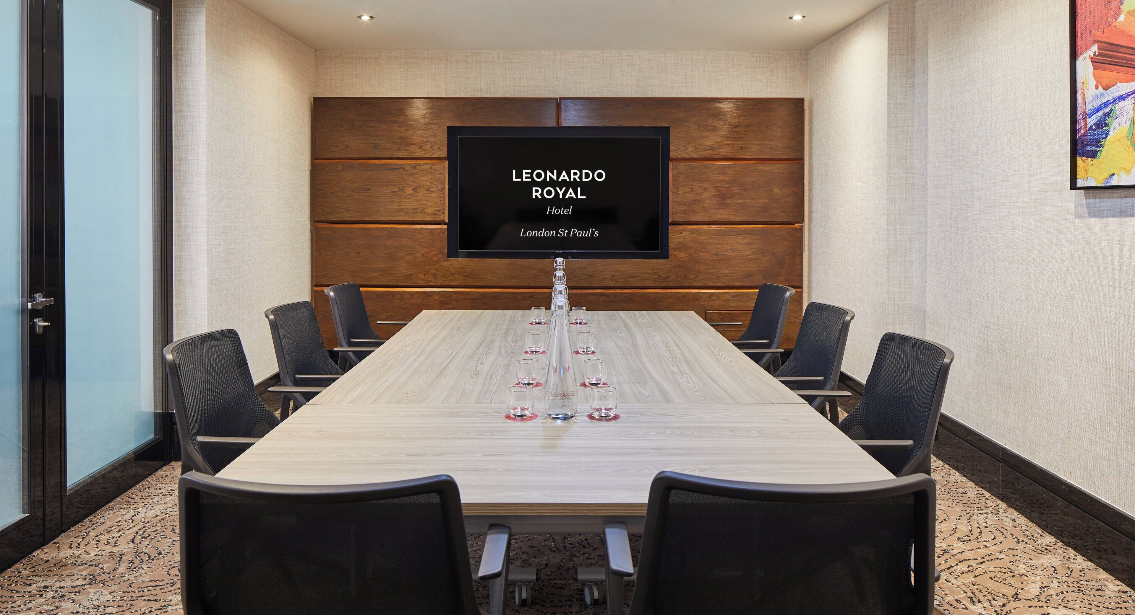 Uma sala de conferência com uma mesa comprida, seis cadeiras e uma tela de televisão exibindo 'LEONARDO ROYAL Hotel London St Paul's'.