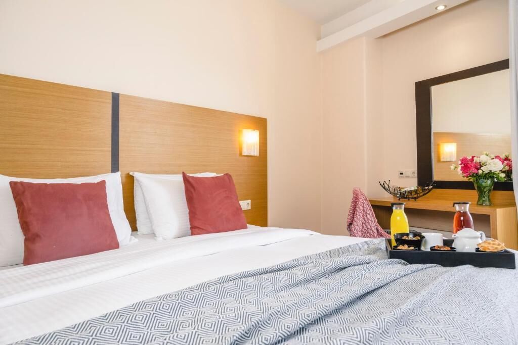 Quarto de hotel com cabeceira de madeira, cama com roupa de cama estampada, mesa de cabeceira com um vaso de flores e bandeja com itens de café da manhã.