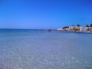 Uma praia com águas claras e rasas, algumas pessoas e um edifício com telhado branco ao fundo.