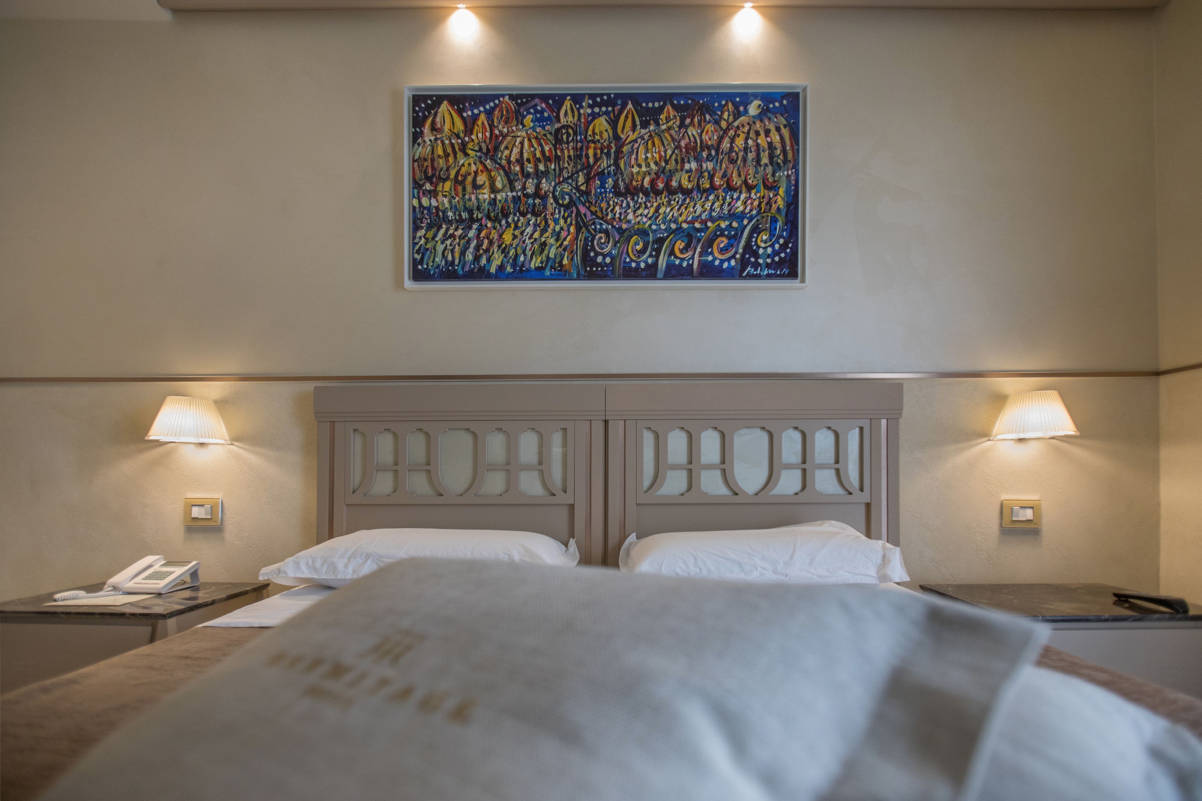 Quarto de hotel com cama, duas luminárias de cabeceira, um telefone na mesinha de apoio e um quadro na parede.
