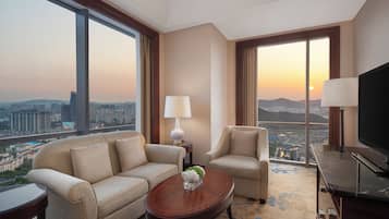 Quarto de hotel com sofá, poltrona, mesa de centro, TV e vista para a paisagem urbana.