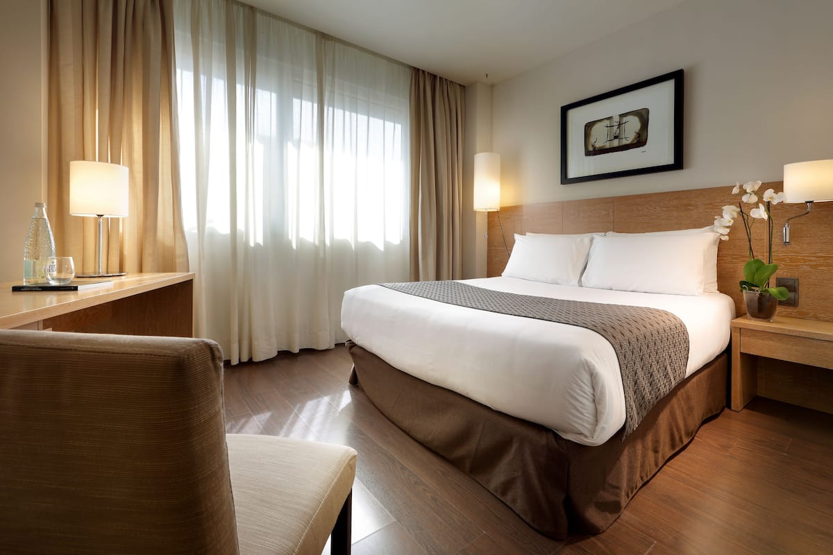 Quarto de hotel com uma cama grande, uma mesa com luminária, uma cadeira e uma mesinha de cabeceira com luminária e planta.