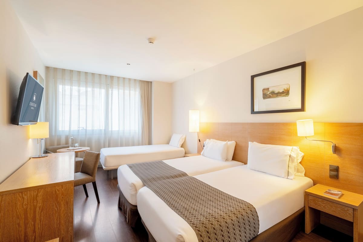 Quarto de hotel com duas camas, uma escrivaninha, uma cadeira, uma TV e um quadro na parede.