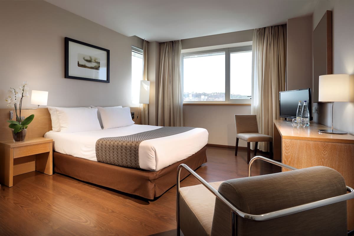 Quarto de hotel com cama, mesas de cabeceira, escrivaninha, cadeira, televisão e janela com cortinas.