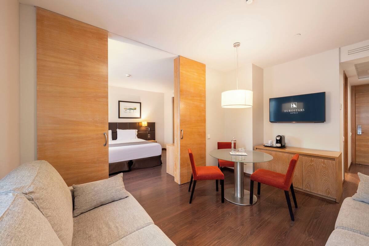 Quarto de hotel com cama, mesa de jantar com cadeiras, televisão fixada na parede e um armário de madeira.