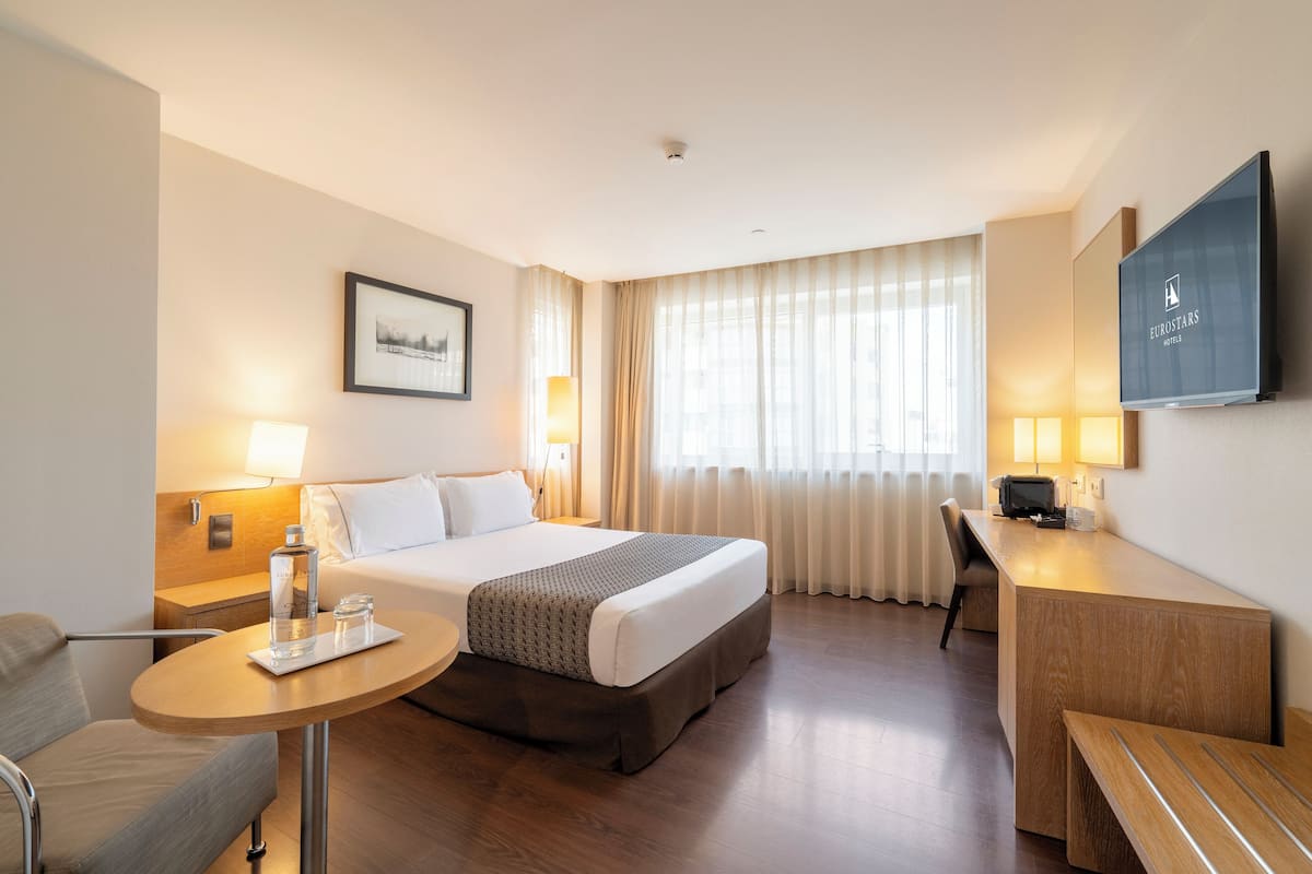 Quarto de hotel com cama, mesas de cabeceira, escrivaninha, cadeira, televisão e janela com cortinas.