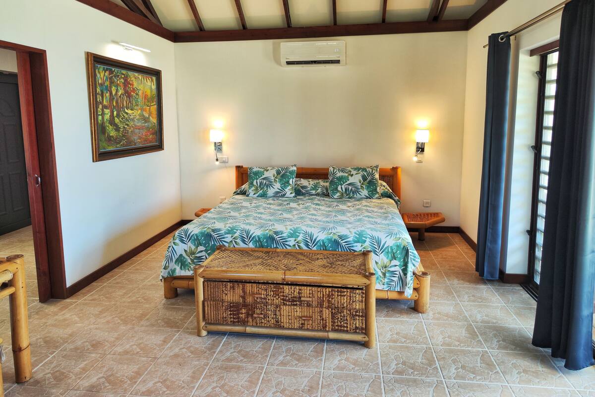 Um quarto com cama, duas mesas de cabeceira, ar-condicionado fixado na parede, um quadro e uma janela com cortinas.