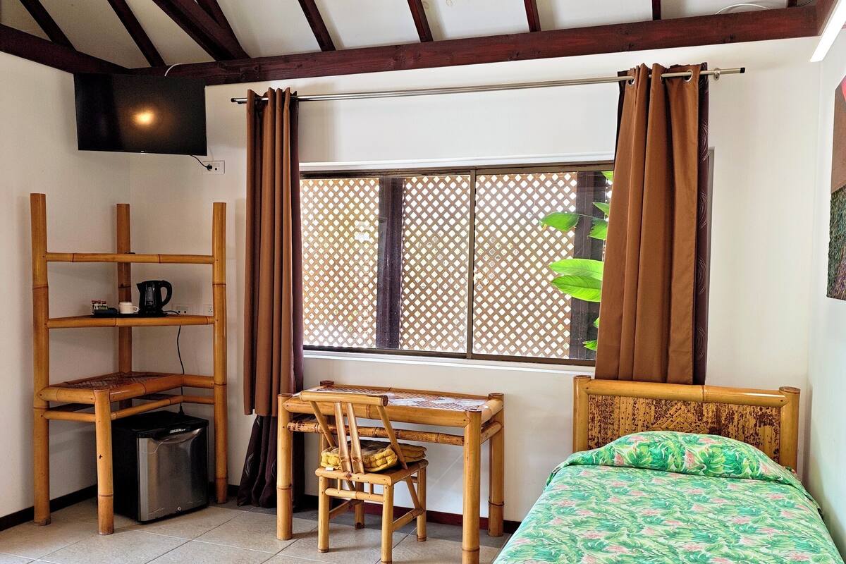 Quarto com cama, escrivaninha, cadeira, televisão e janela com cortinas.
