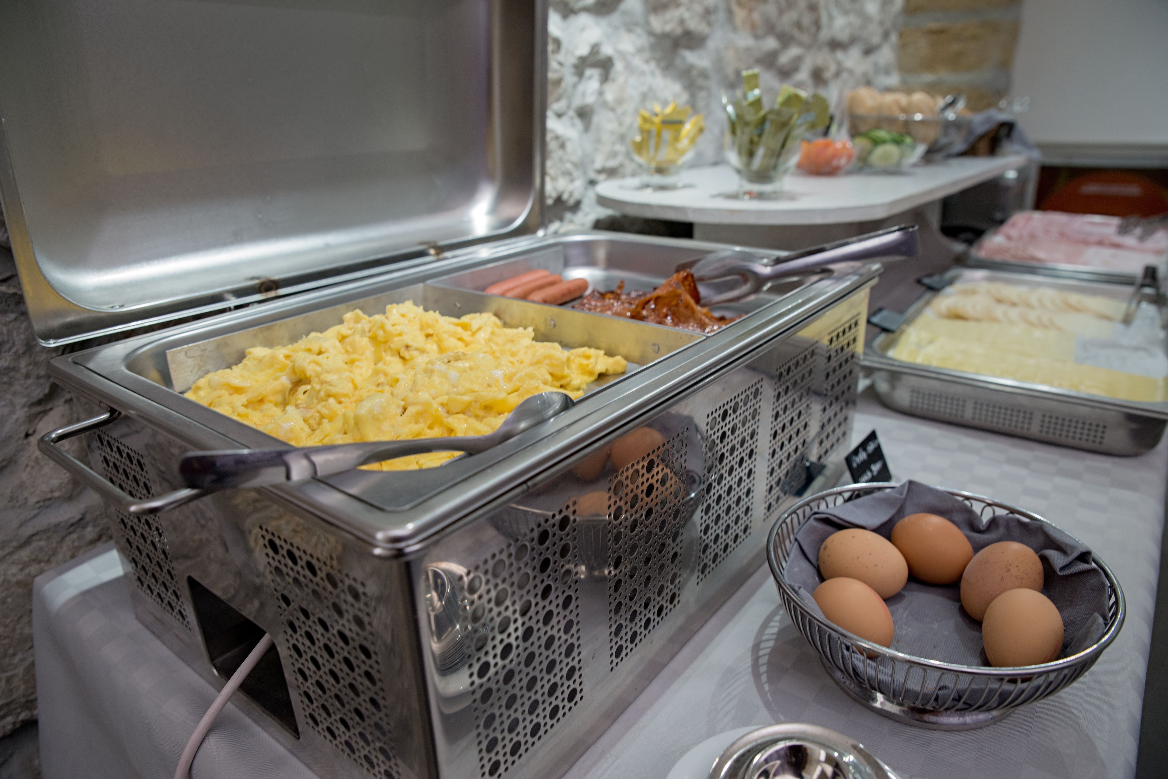 Buffet com diversos alimentos, incluindo ovos mexidos, salsichas e ovos em uma tigela.