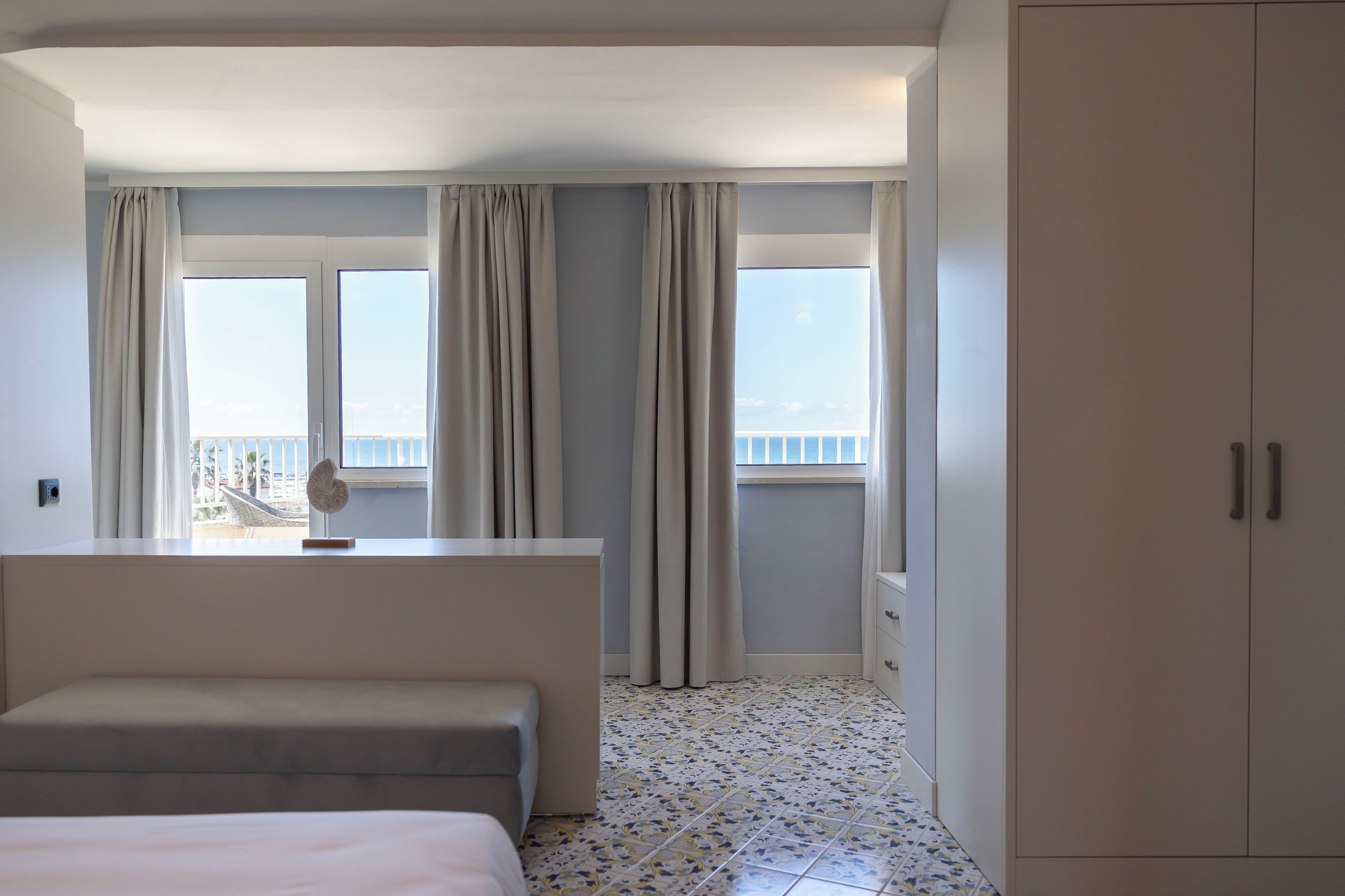 Quarto de hotel com cama, escrivaninha, armário e amplas janelas com cortinas.