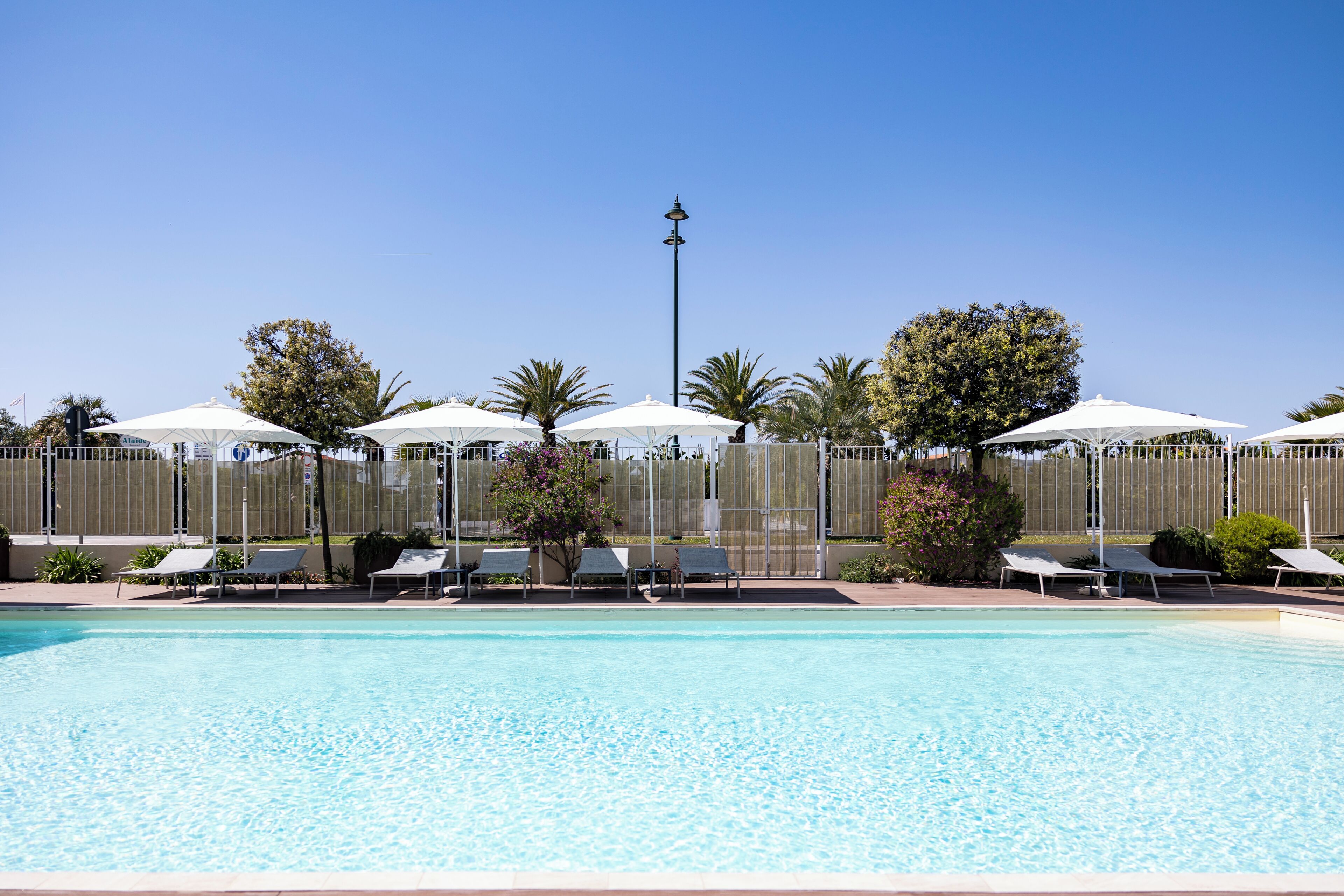 Piscina com espreguiçadeiras e guarda-sóis, cercada por uma grade e árvores.