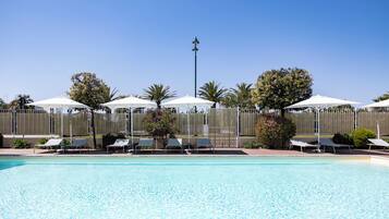 Piscina com espreguiçadeiras e guarda-sóis, cercada por uma grade e árvores.