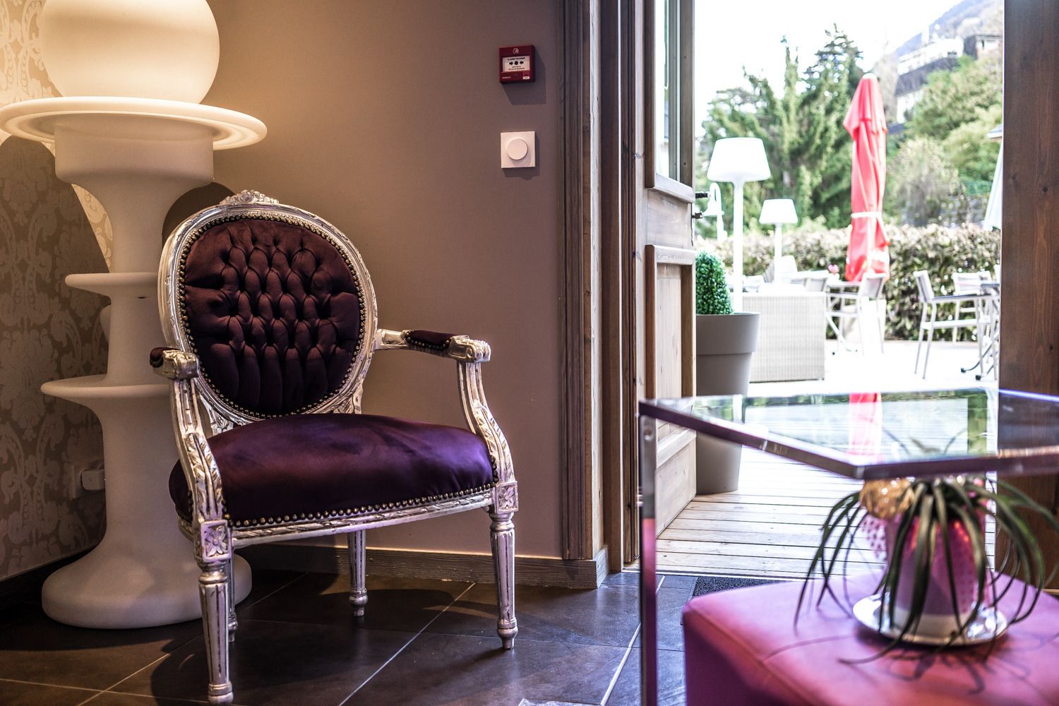 Uma cadeira luxuosa com encosto alto e estofamento macio, posicionada ao lado de um abajur branco sobre um tampo de vidro com uma almofada rosa.