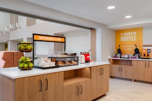 Uma estação de café da manhã com um letreiro que diz 'CAFÉ DA MANHÃ FELIZ COMEÇA AQUI' e uma estação de waffles com um letreiro que diz 'THE HAMPTON WAFFLE'.
