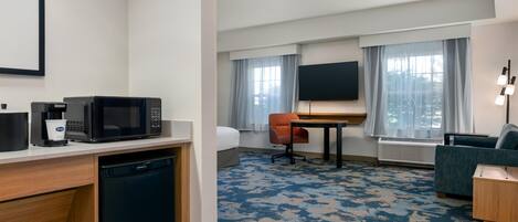 Quarto de hotel com cozinha compacta, cama, escrivaninha, cadeira e televisão.