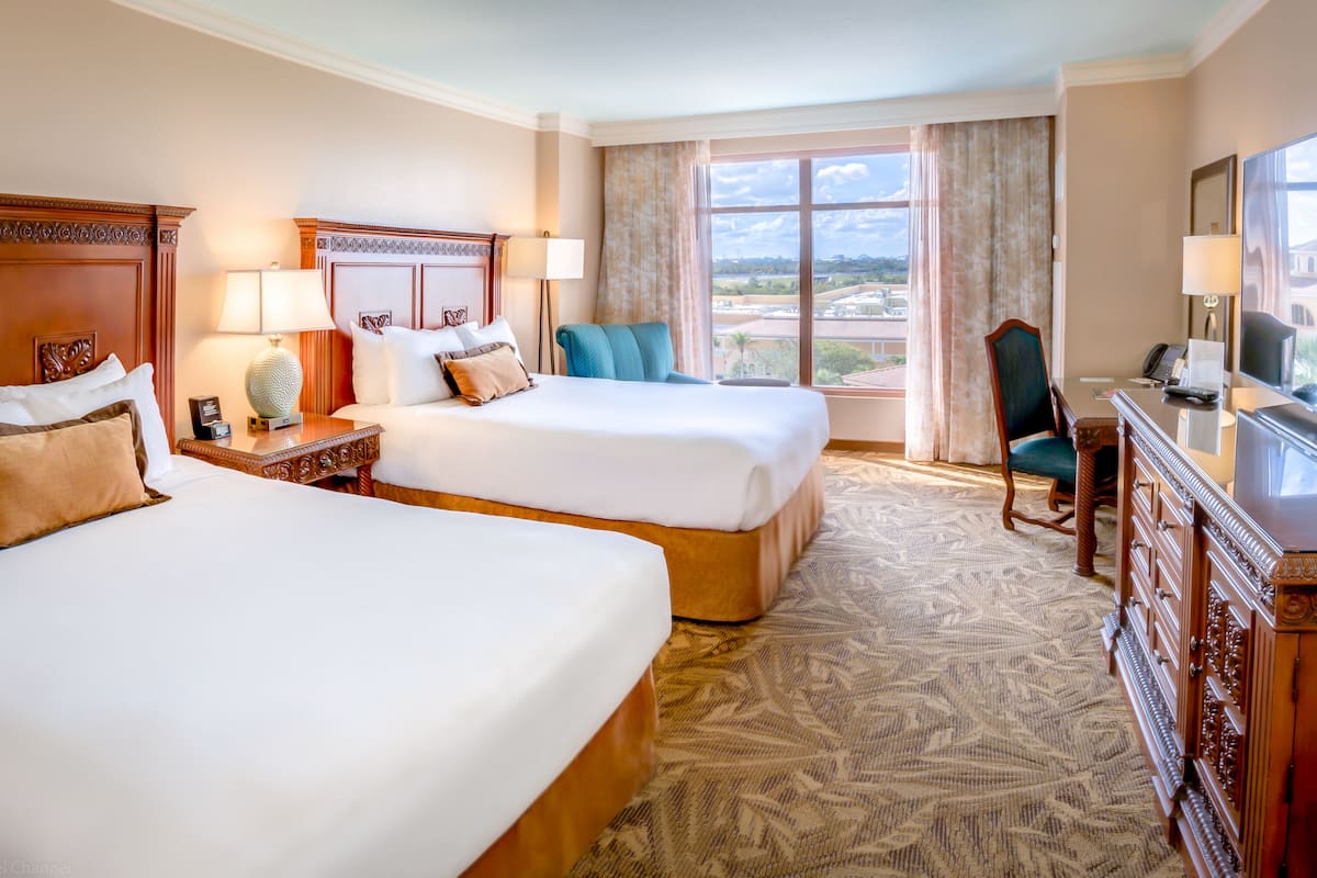Quarto de hotel com duas camas, uma escrivaninha, uma cadeira, uma TV e uma grande janela com vista.