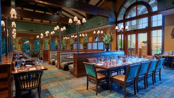Uma ampla sala de jantar com várias mesas e cadeiras, janelas grandes e plantas decorativas.
