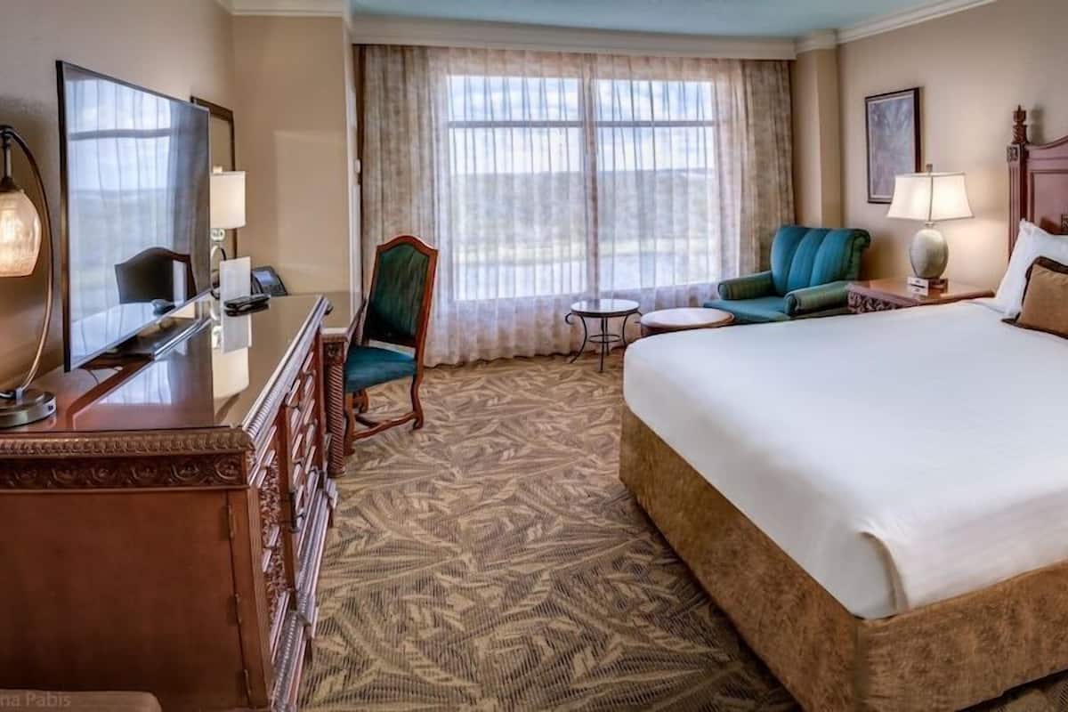 Quarto de hotel com uma cama grande, uma escrivaninha com abajur, uma cadeira, uma mesinha e uma janela com cortinas.