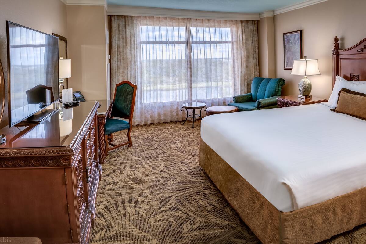Quarto de hotel com uma cama grande, uma escrivaninha com telefone, uma cadeira, uma mesinha e uma janela com cortinas.